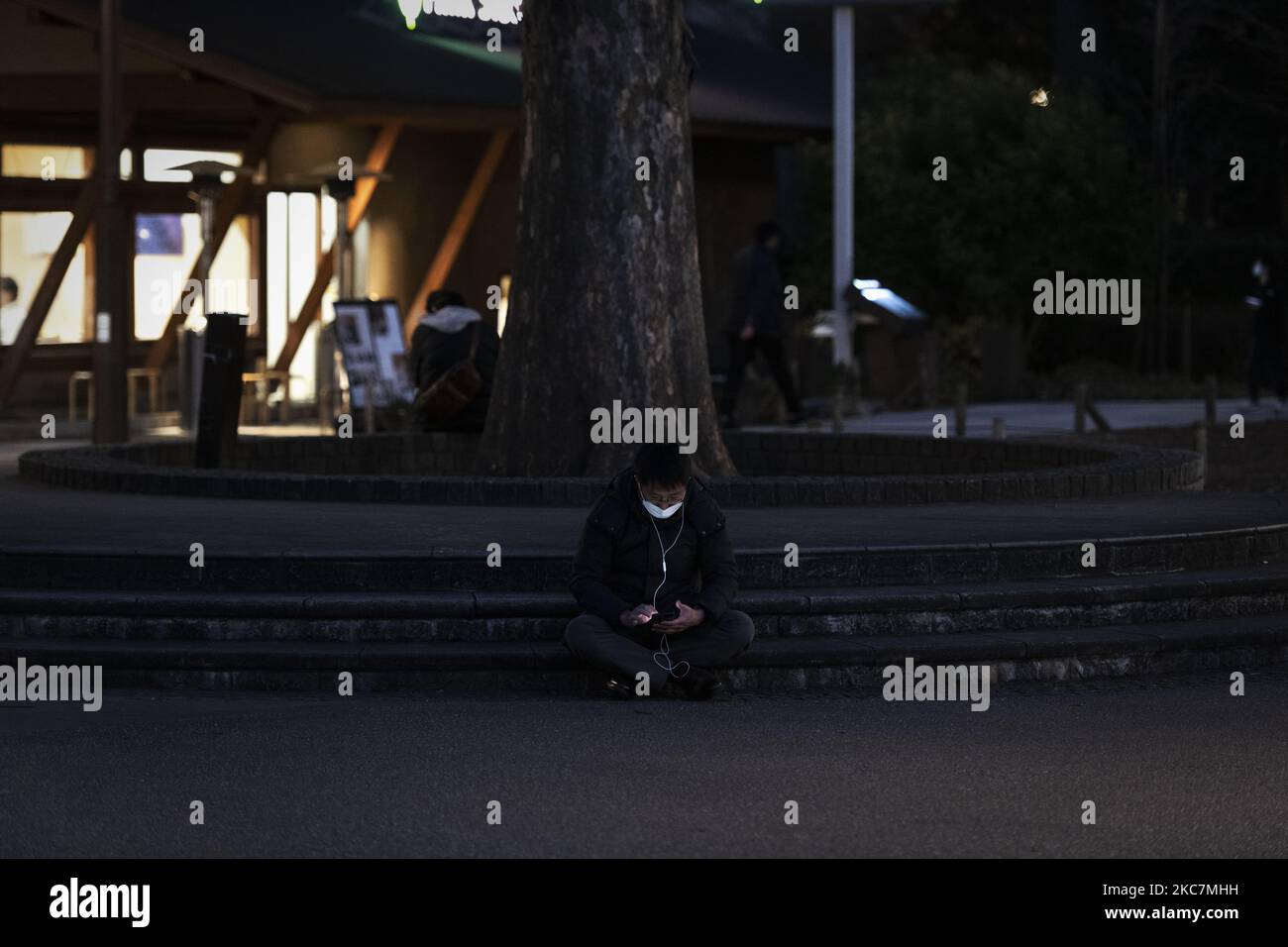 Man wearing a protective face mask plays his mobile at dusk in the Ueno ...