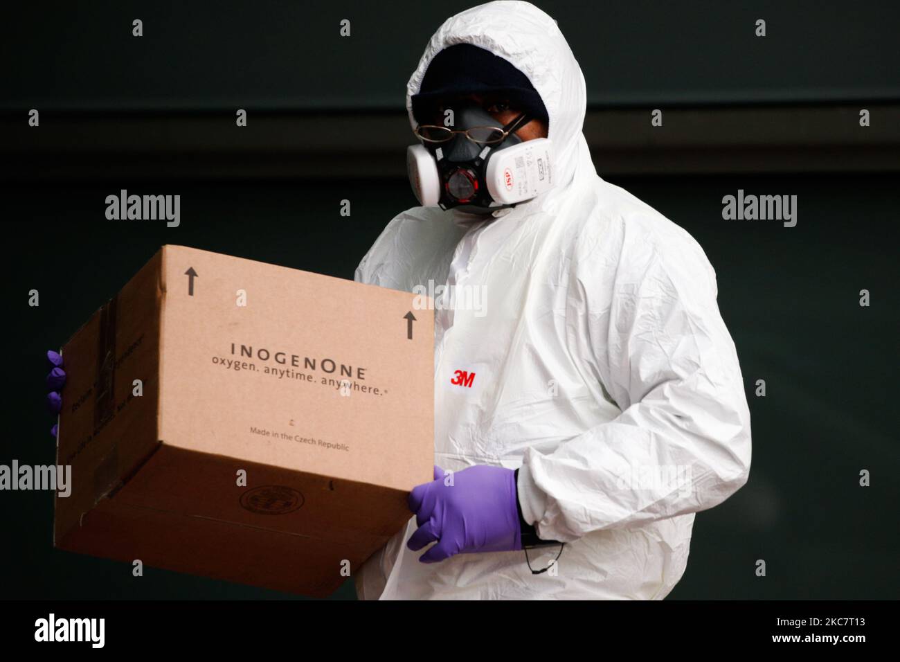 A delivery man wearing a PPE suit and respirator mask carries a package ...