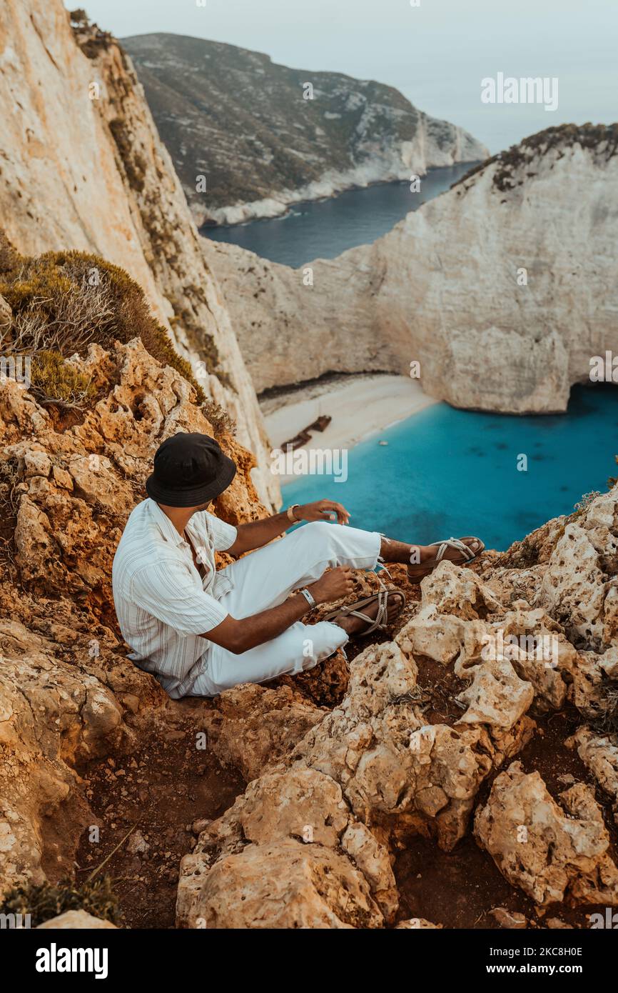 A back view of black man sitting on steep cliff overlooking the canyon ...