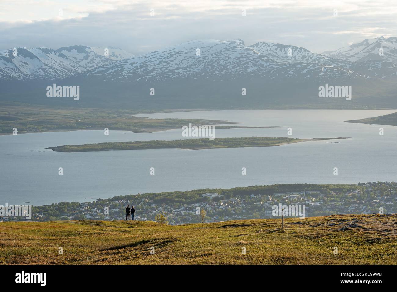 A scenic view of a couple on a green hill against a lake and a mountain ...