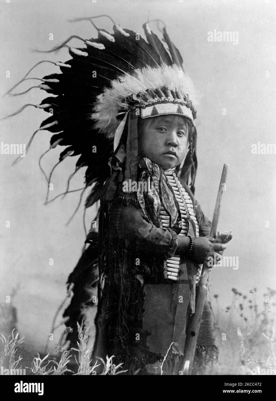 A young Native American boy sporting a full headdress and holding a ...