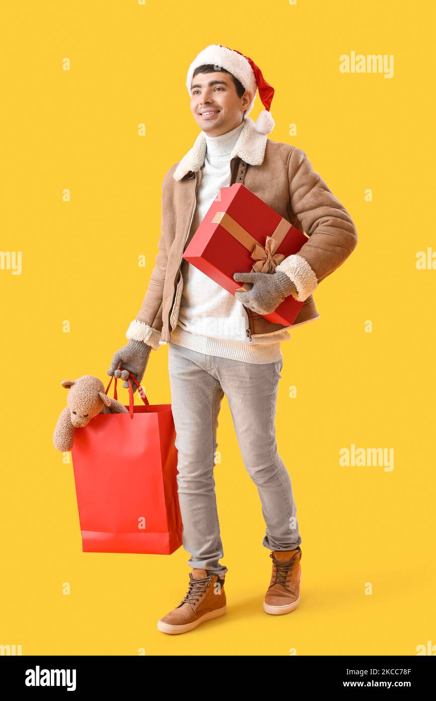 Young man in Santa hat with Christmas gift and shopping bag on yellow ...
