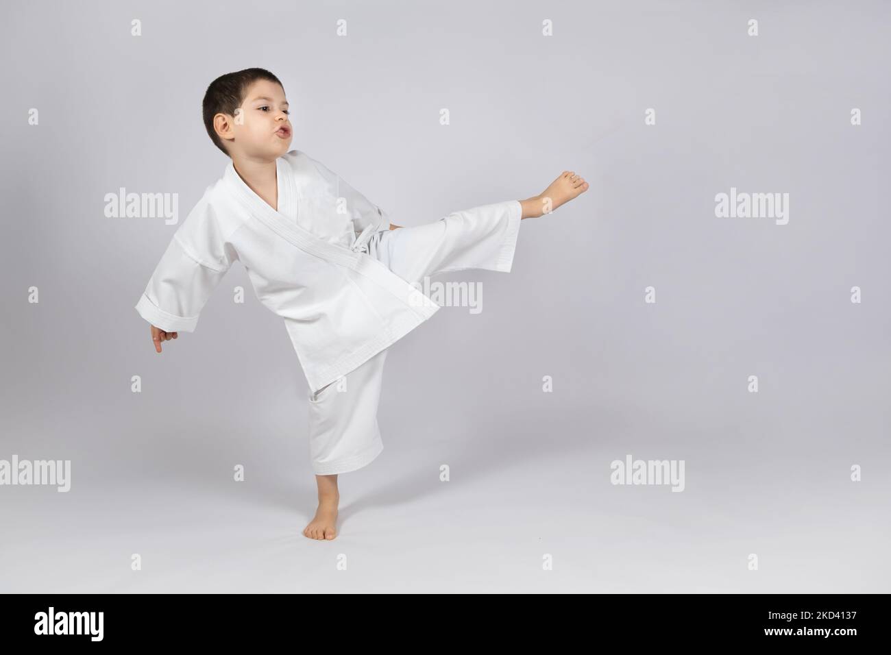 A little boy in a kimono practices karate on a white background ...