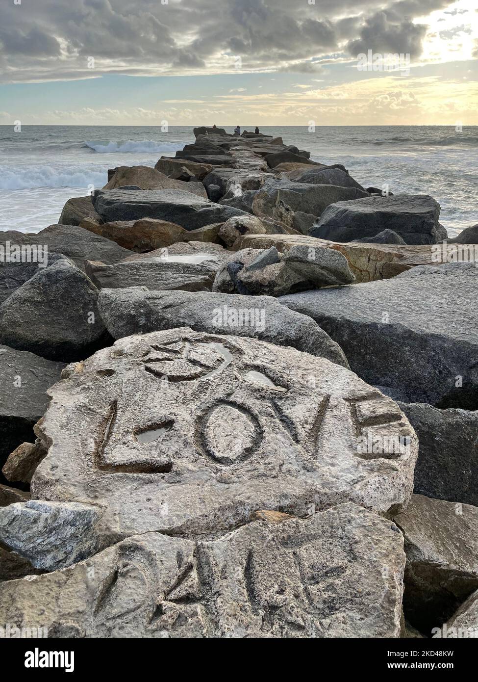 A vertical closeup of a stone line carved the text "love", sea waves ...