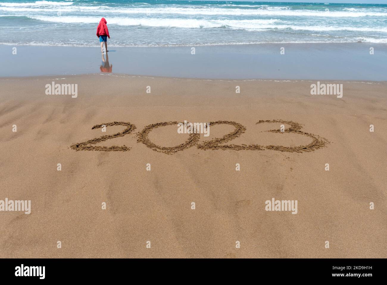 A year 2023 written in the sand on the beach with a boy in a red coat ...