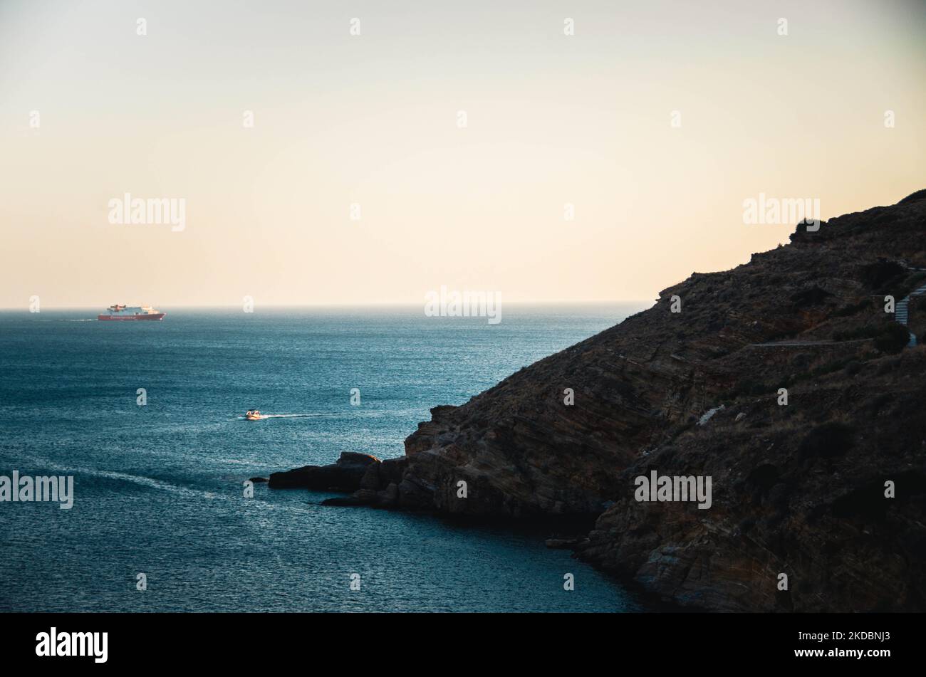 A vast sea stack with boats sailing on the blue water below in Ios ...