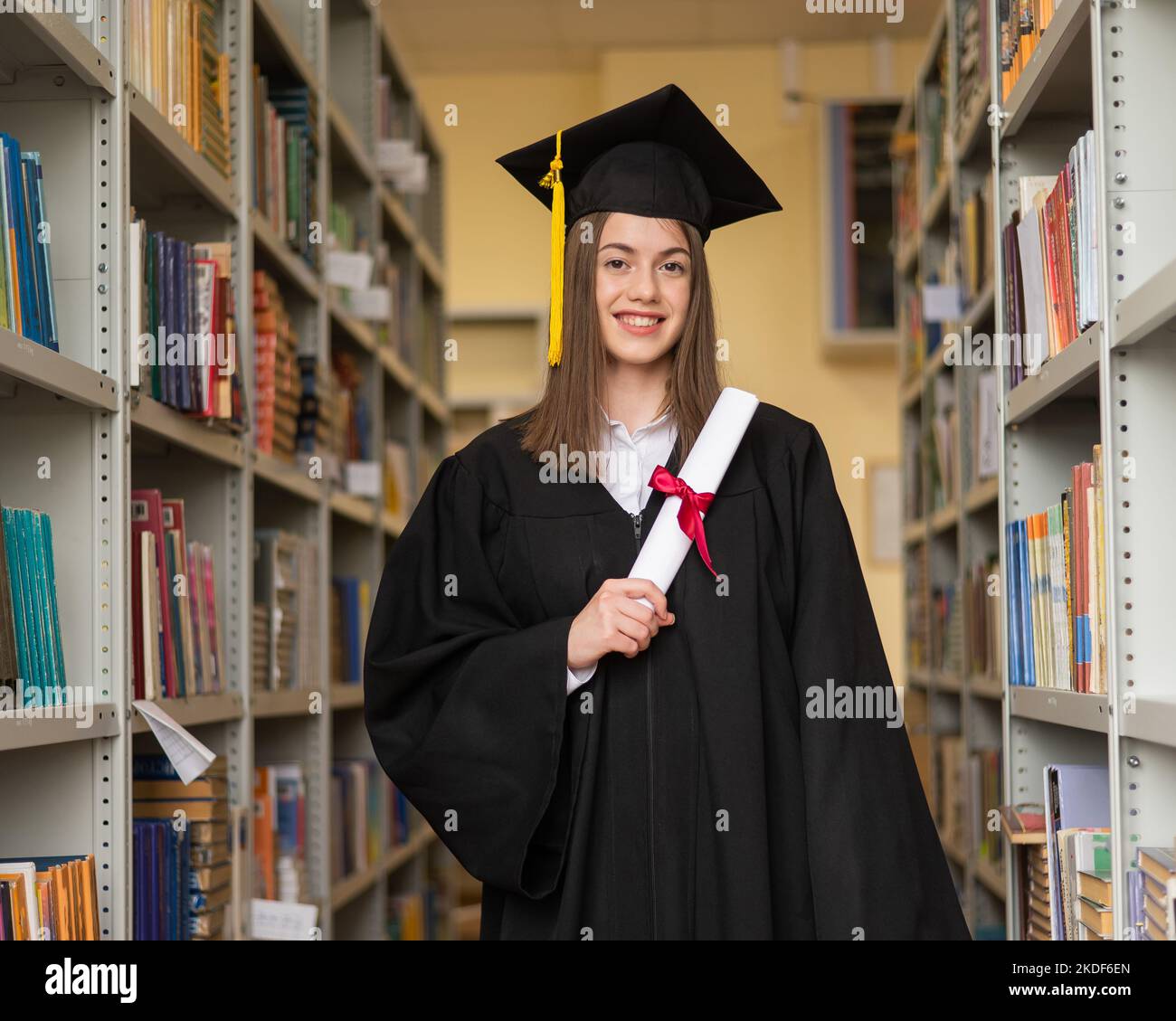 Happy young woman in graduate gown holding diploma in the library Stock ...