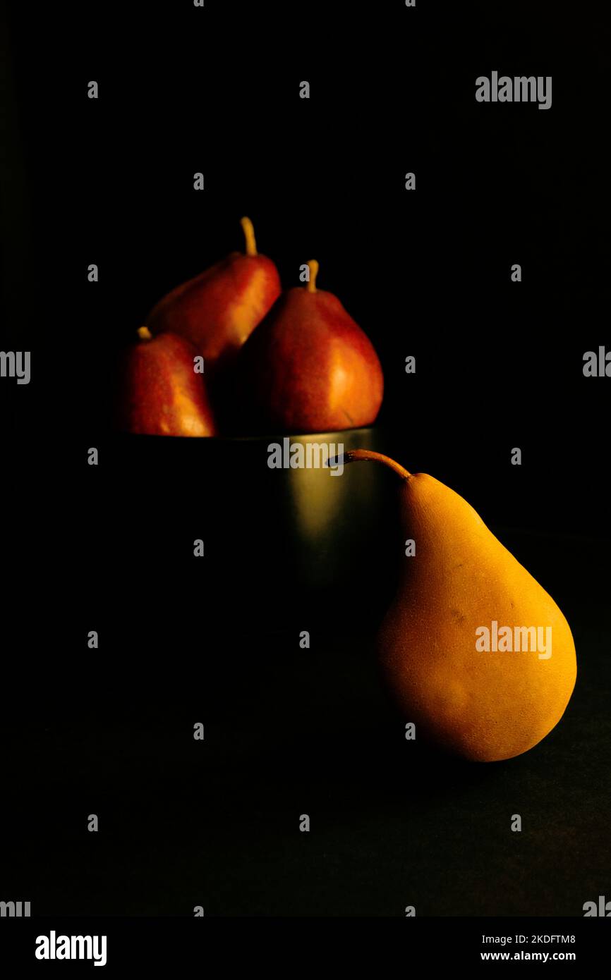 A yellow pear in front of a dark bowl full of red pears on dark ...