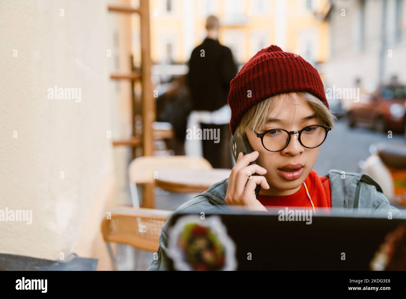 Asian boy talking on mobile phone while working with laptop in cafe ...
