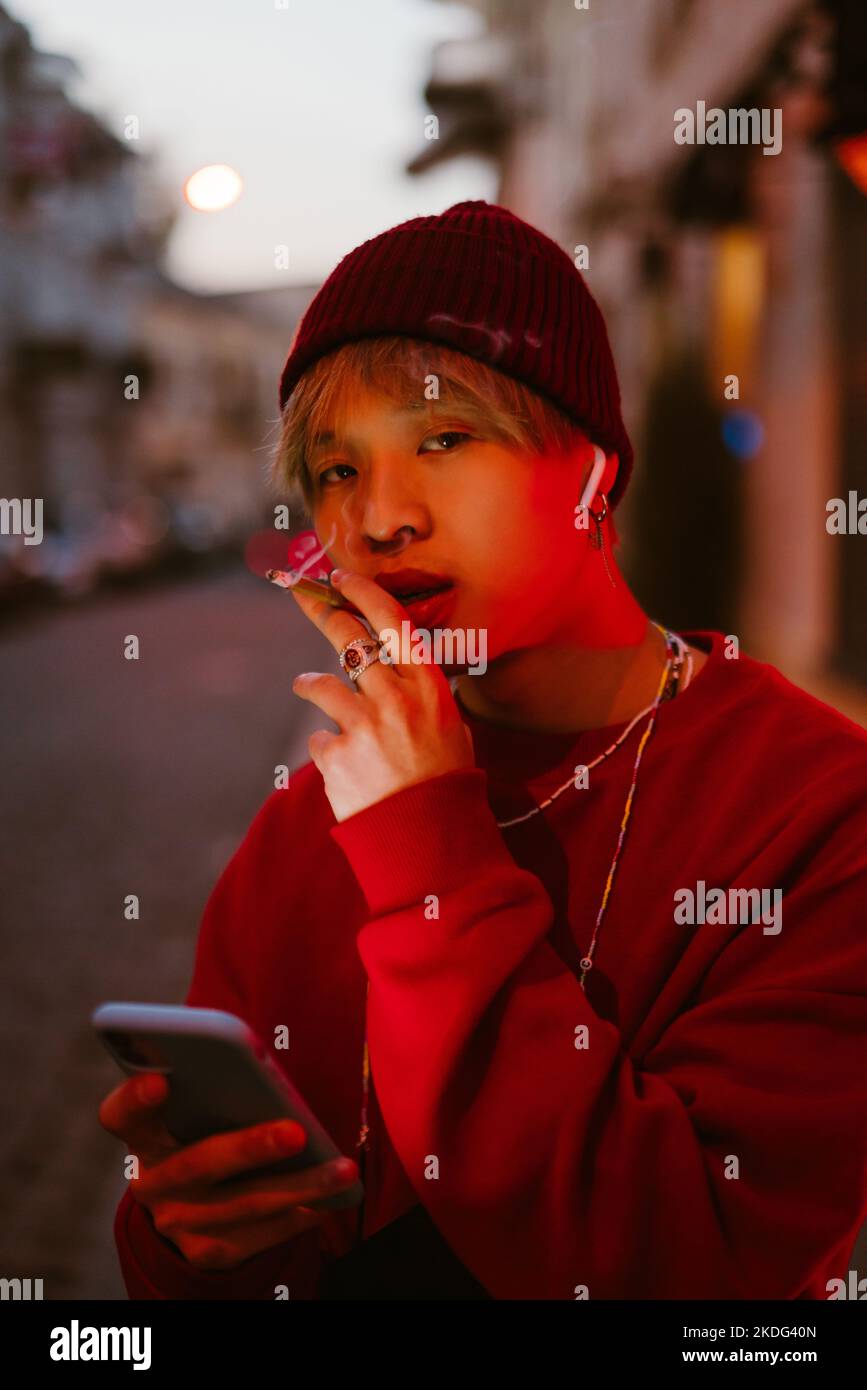 Asian boy in earphones using mobile phone while smoking at city street ...