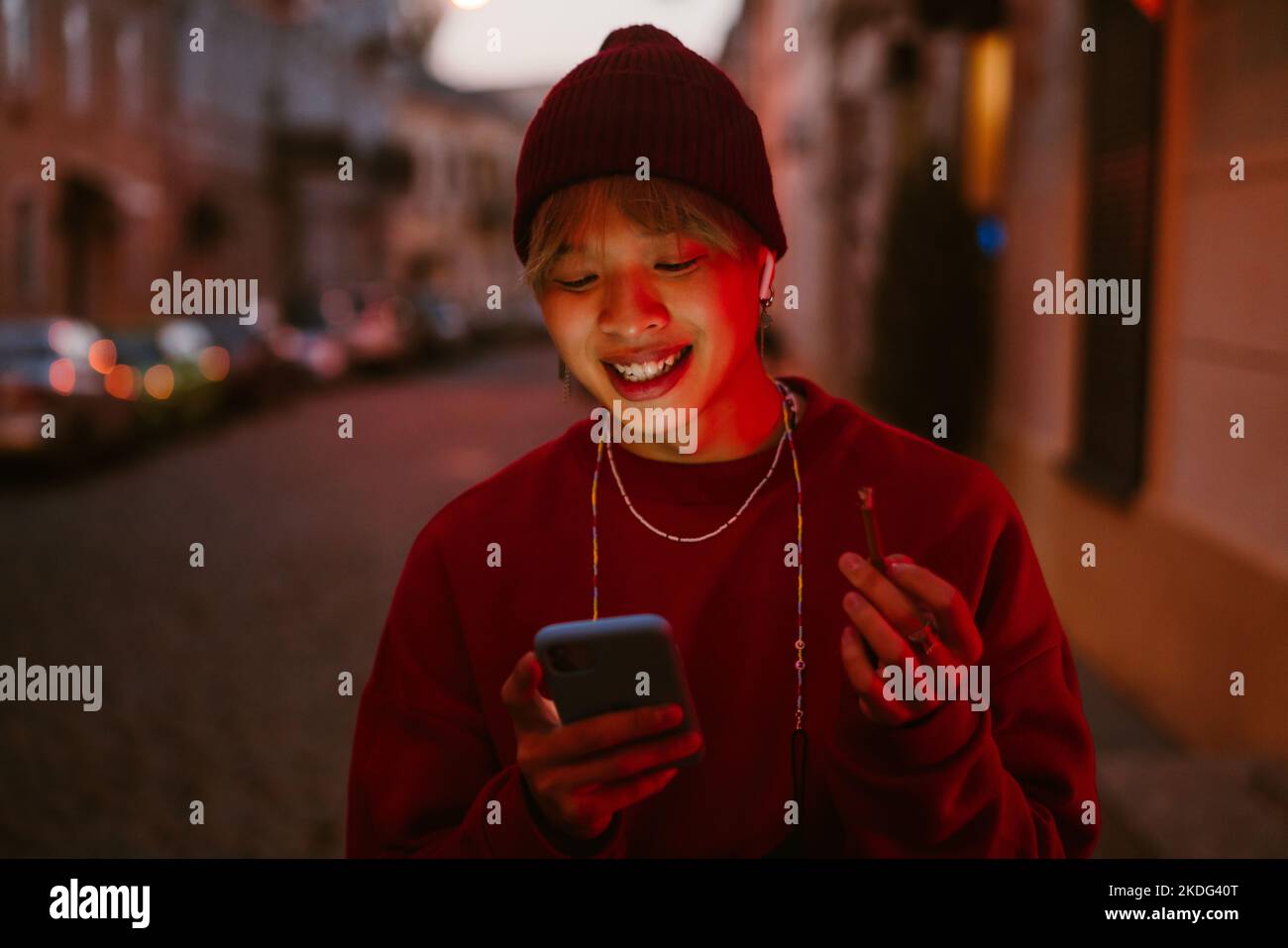 Asian boy in earphones using mobile phone while smoking at city street ...