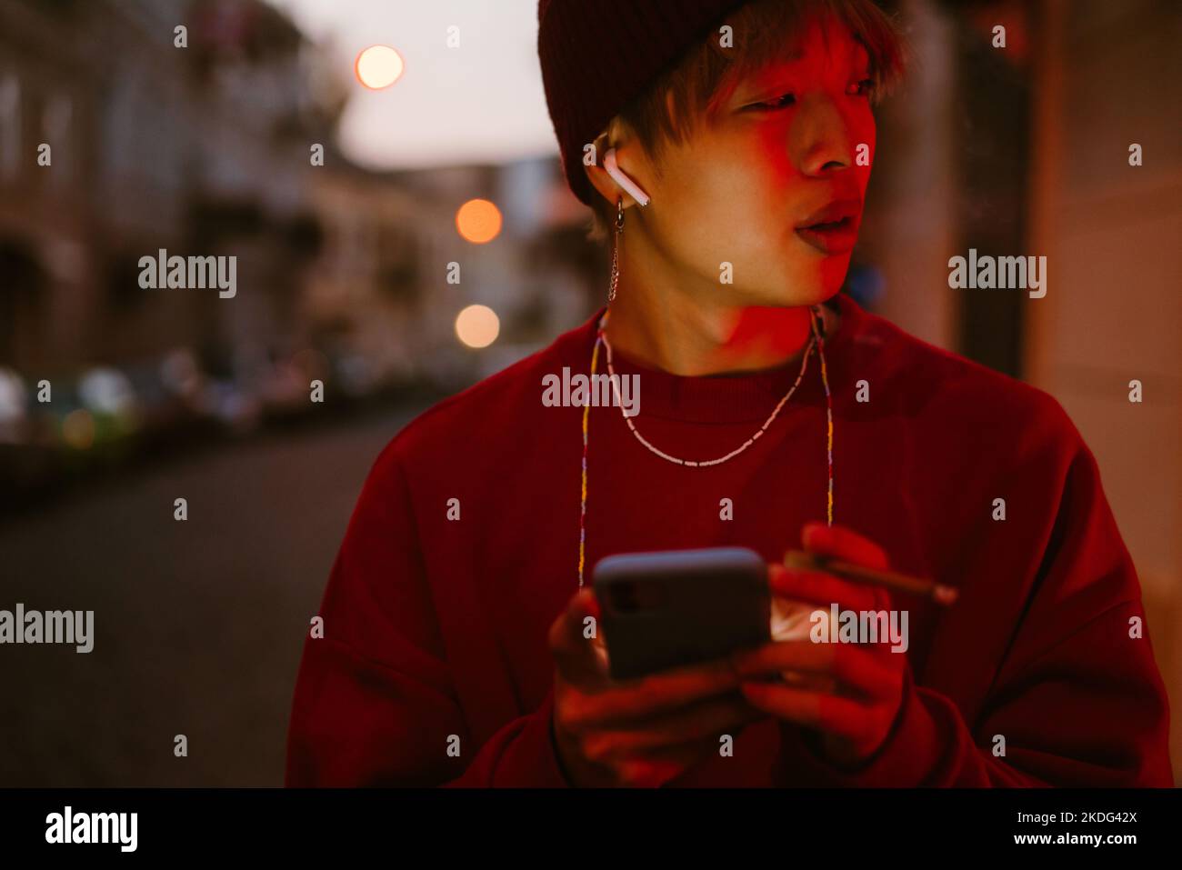 Asian boy in earphones using mobile phone while smoking at city street ...