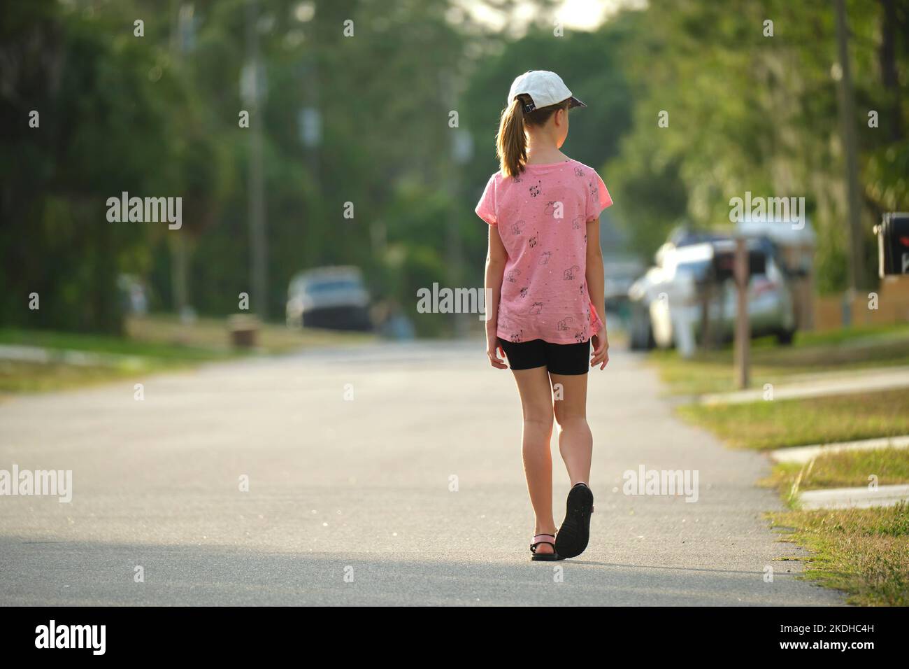 Rear view of confident young child girl walking along the sunny alley ...