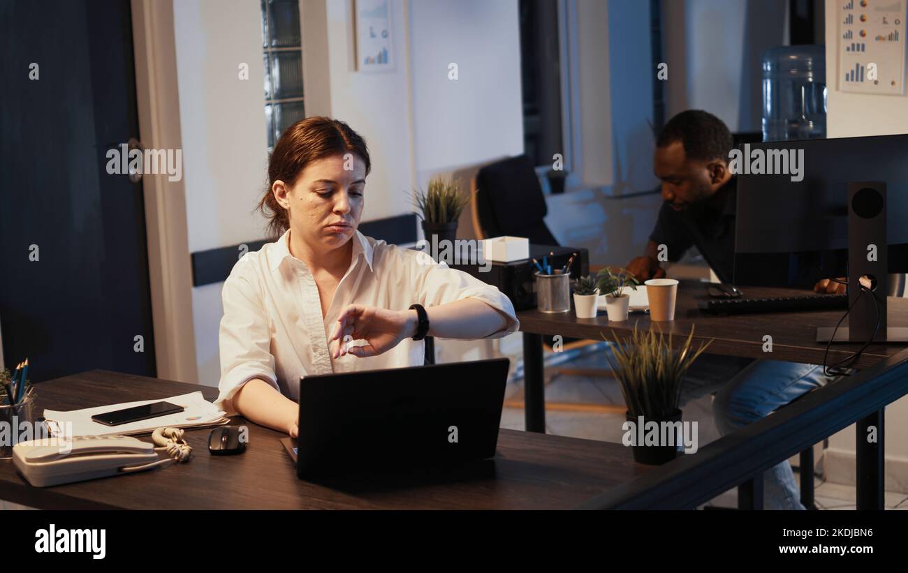 Office employee working on laptop at night and checking time on ...