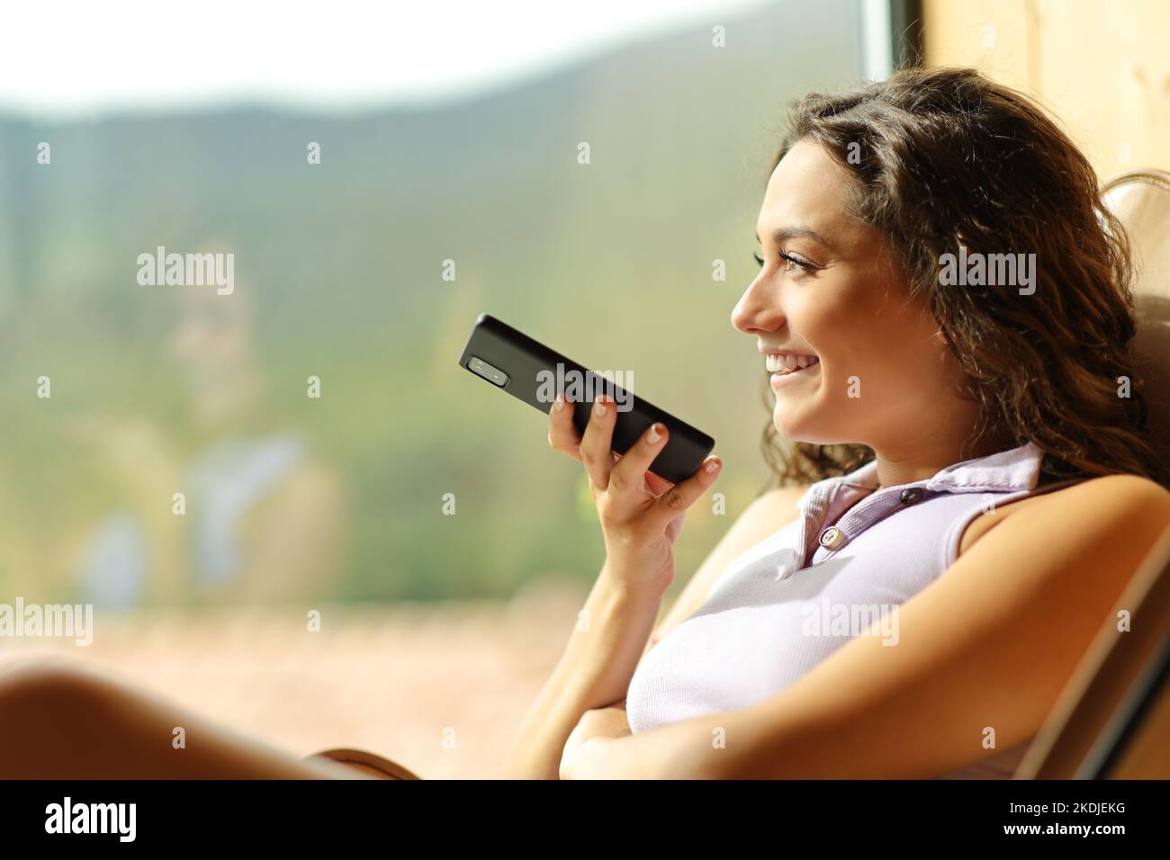 Happy woman sitting on a chair dictating message on smart phone looking ...