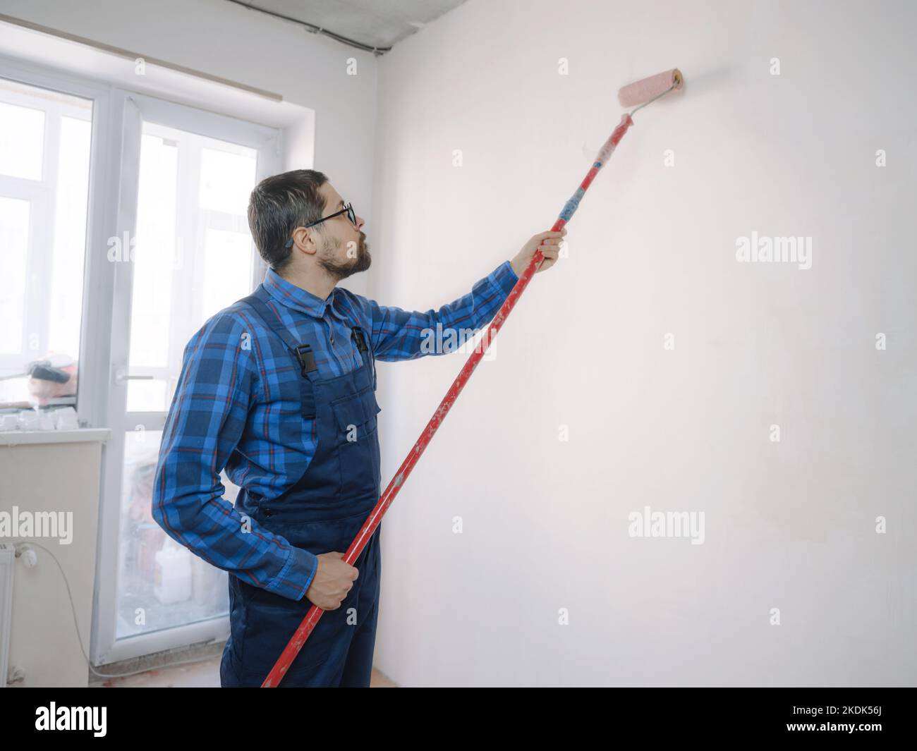young man in blue work suit hold paint roller, over white wall room ...