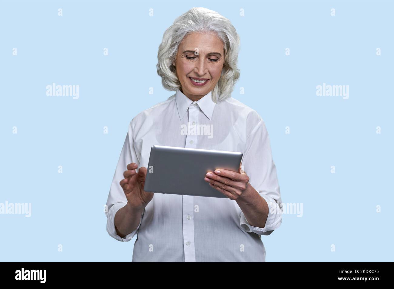 Smiling senior gray-haired woman using tablet pc. Isolated on pale blue ...