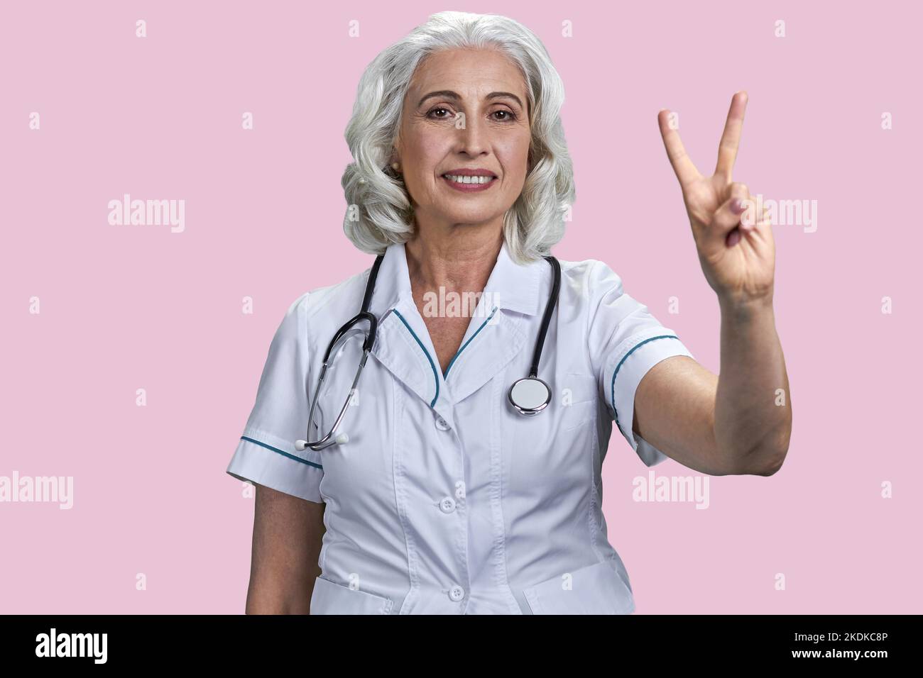 Aged caucasian female doctor show victory sign. Isolated on pink ...