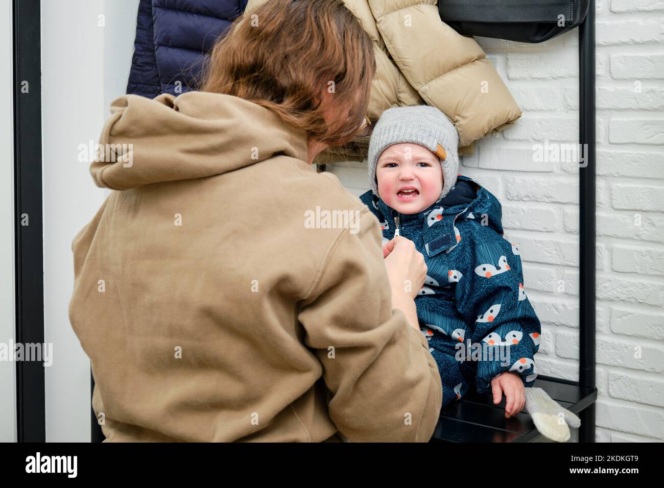 Mother puts a overalls on the cry toddler baby sitting in the home ...