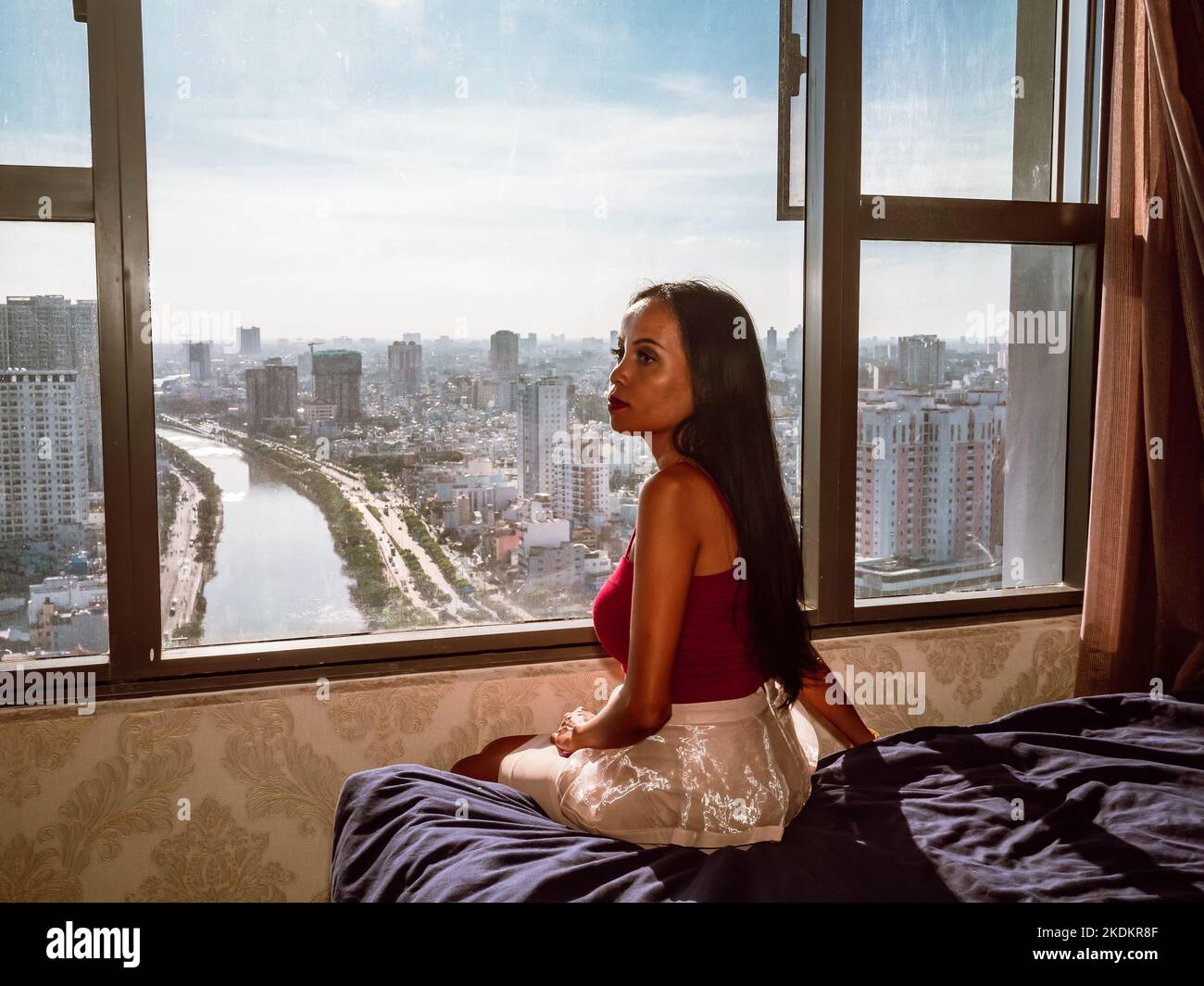 Side view of an Asian woman sitting on a bed at the window with the ...