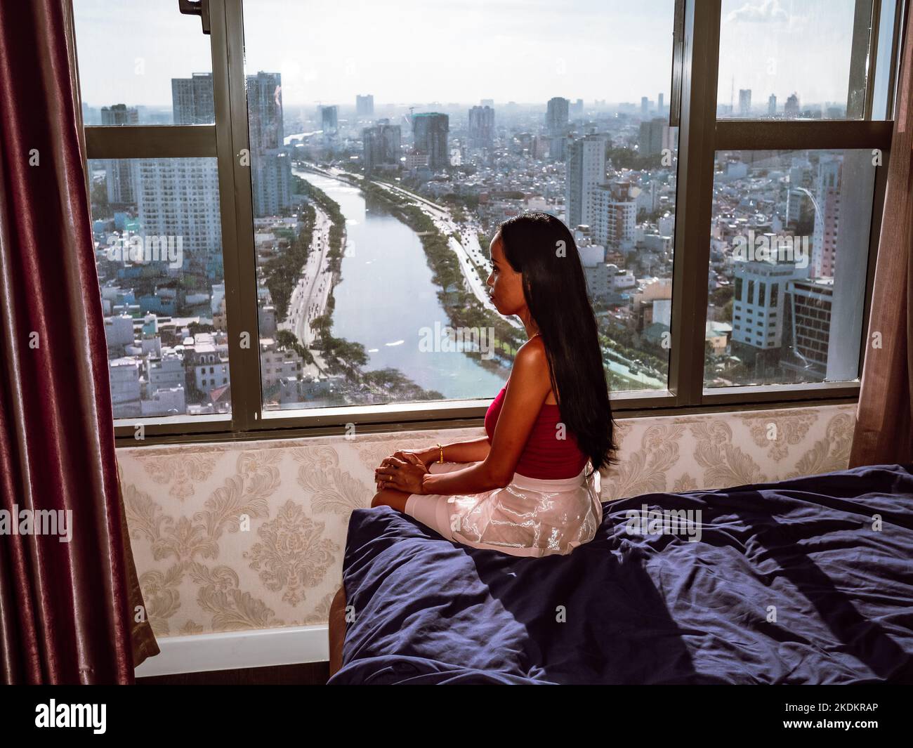 Side view of an Asian woman sitting on a bed at the window with the ...