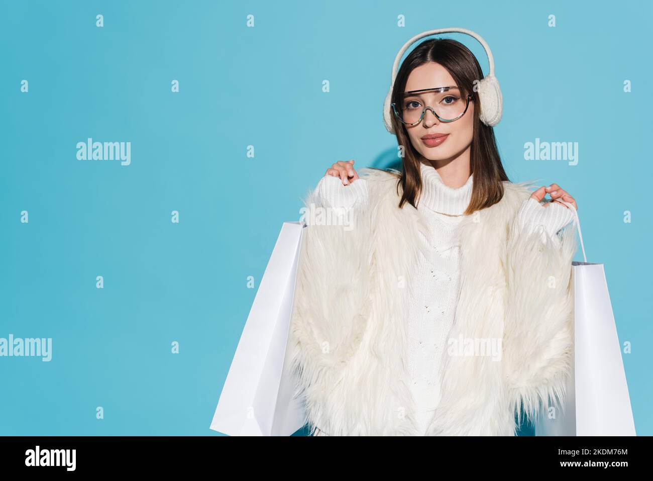 young woman in earmuffs and stylish faux fur jacket holding shopping ...