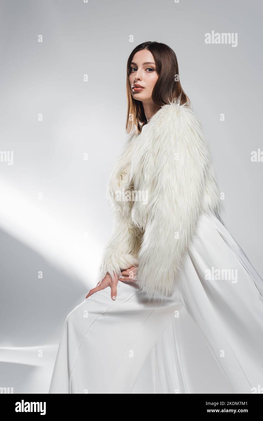 Trendy model in white clothes and faux fur jacket looking at camera on ...