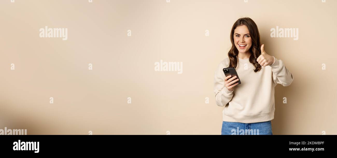 Young woman smiling, showing thumbs up while using mobile phone ...