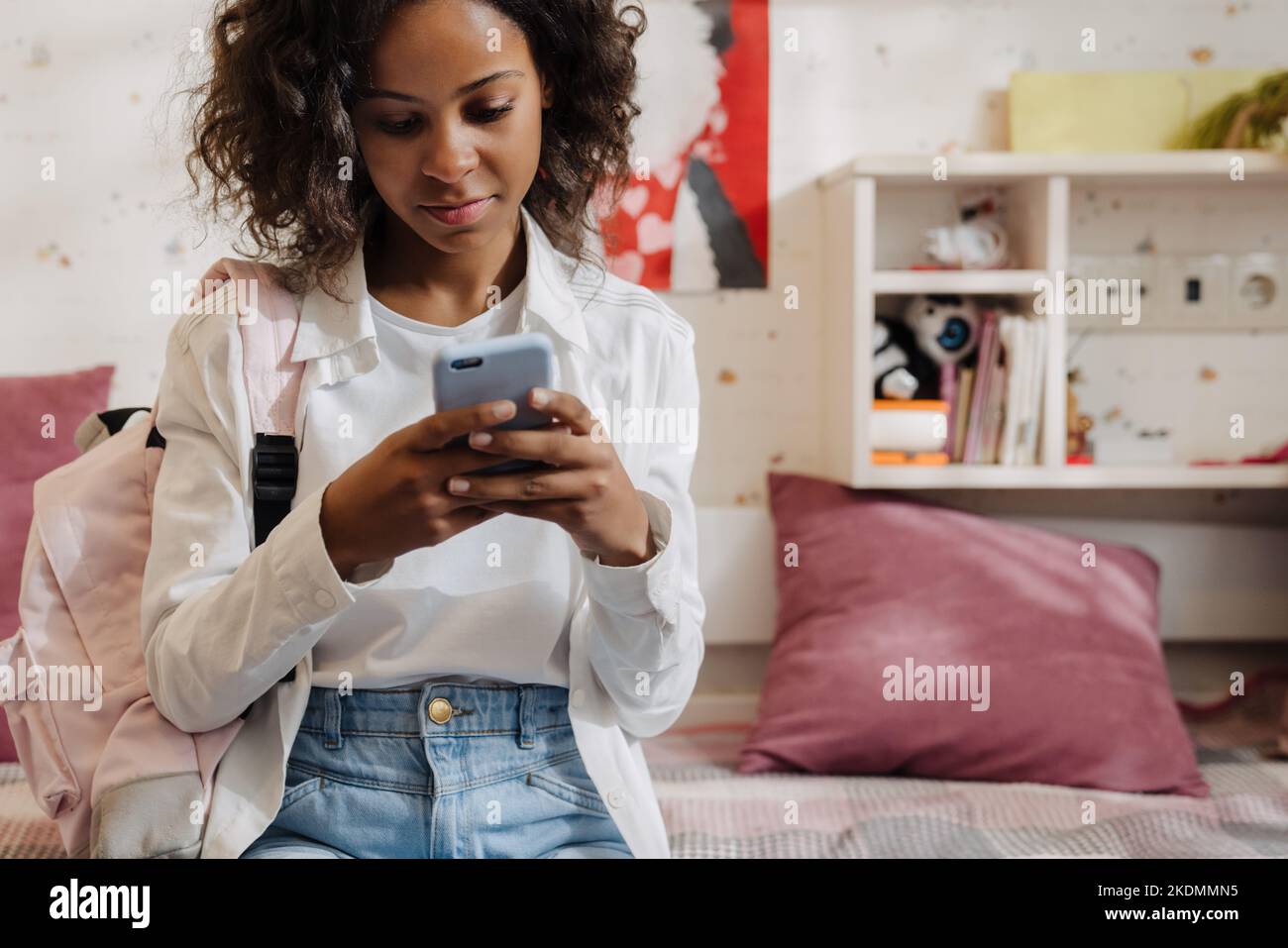 African calm teen girl sitting on a bed with phone texting or using ...