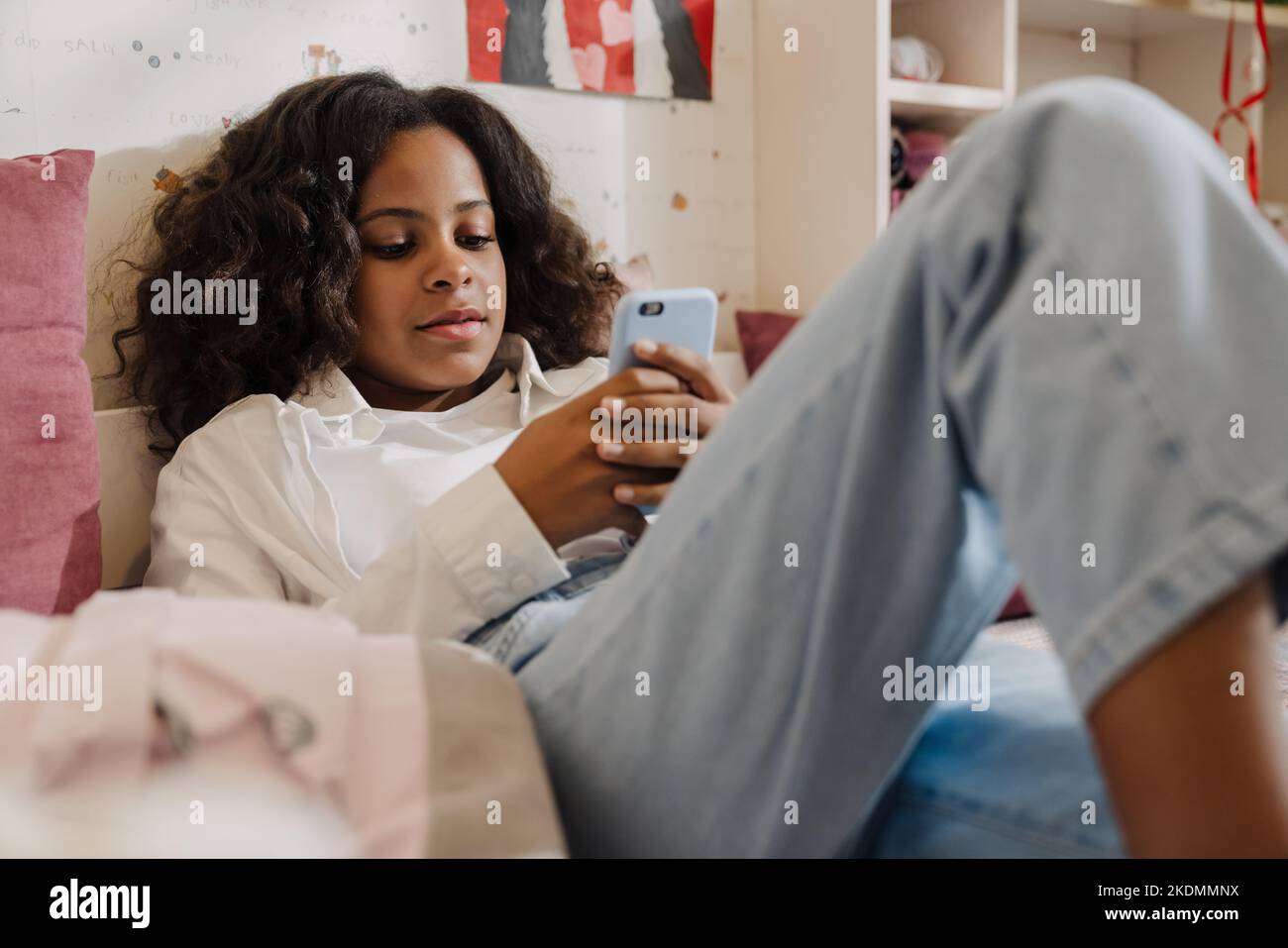 African calm teen girl liying on a bed with phone texting or scrolling ...