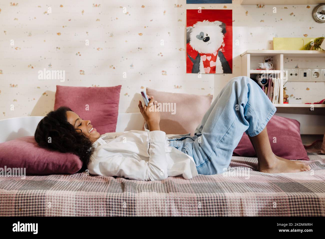 African smiling teen girl liying on a bed with phone surfing on ...