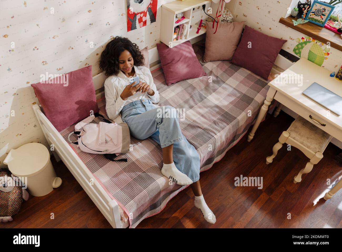 African calm teen girl sitting on a bed with phone after school in her ...