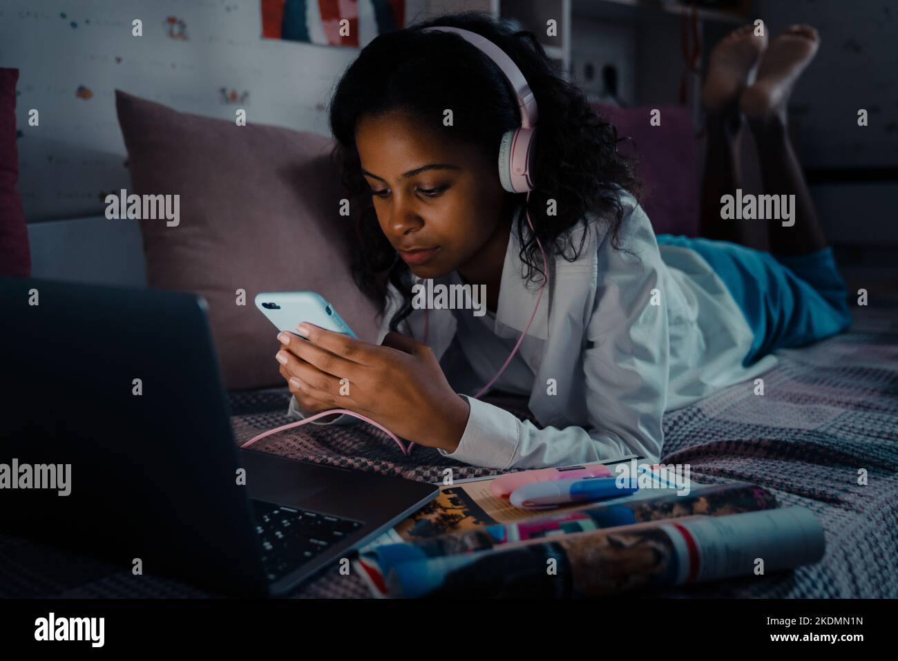 African calm teen girl in headphones liying on a bed with phone texting ...