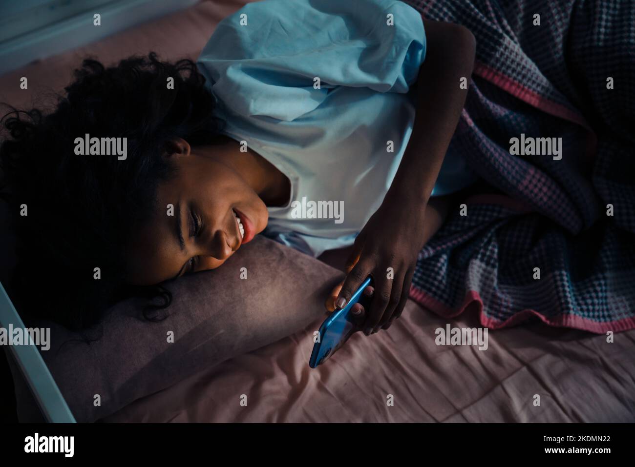 African smiling beautiful teen girl using phone before sleep in dark ...