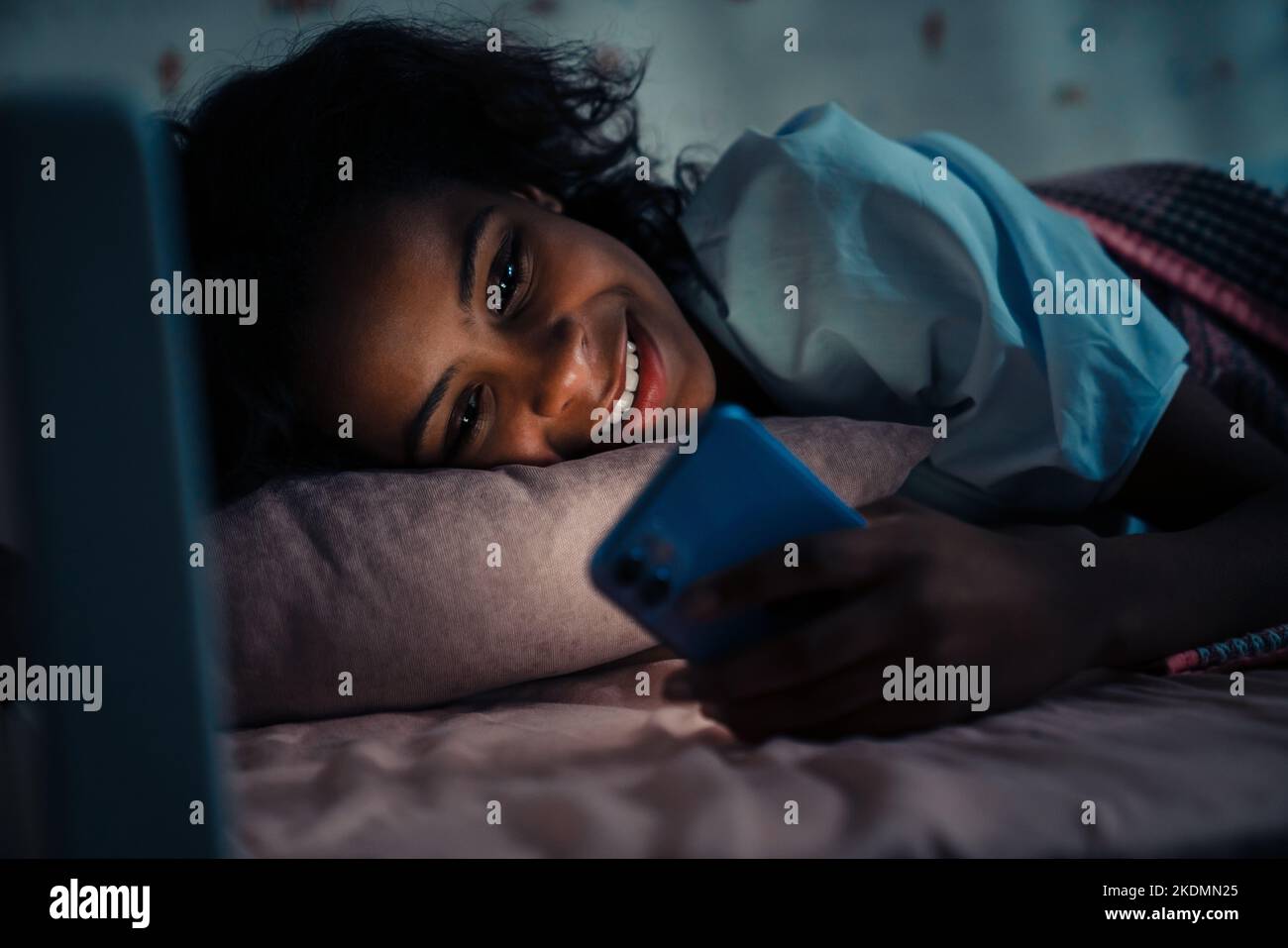 African smiling beautiful teen girl using phone before sleep in dark ...