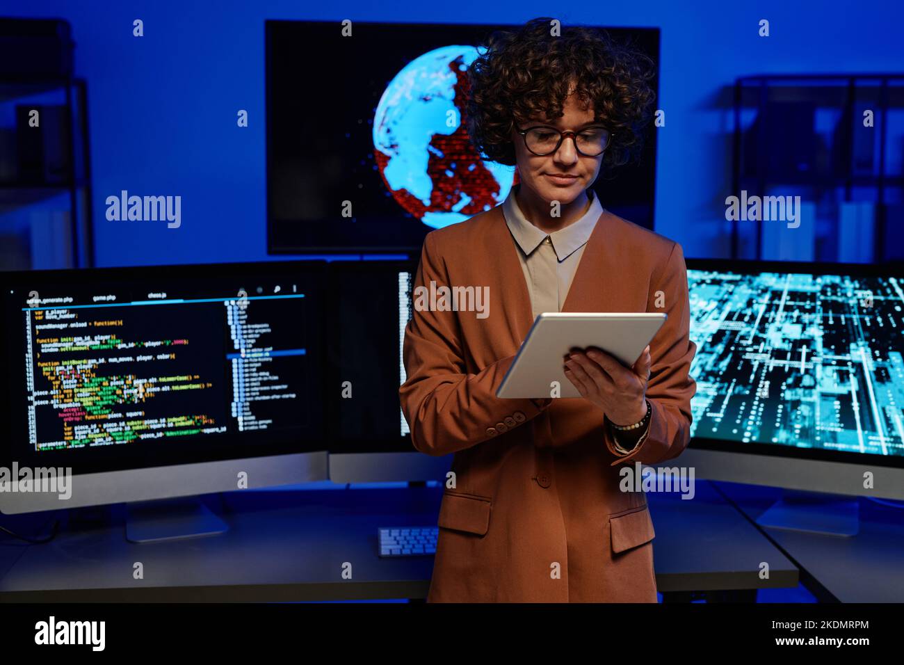 Young serious female programmer using tablet while standing against computer monitors with coded ...