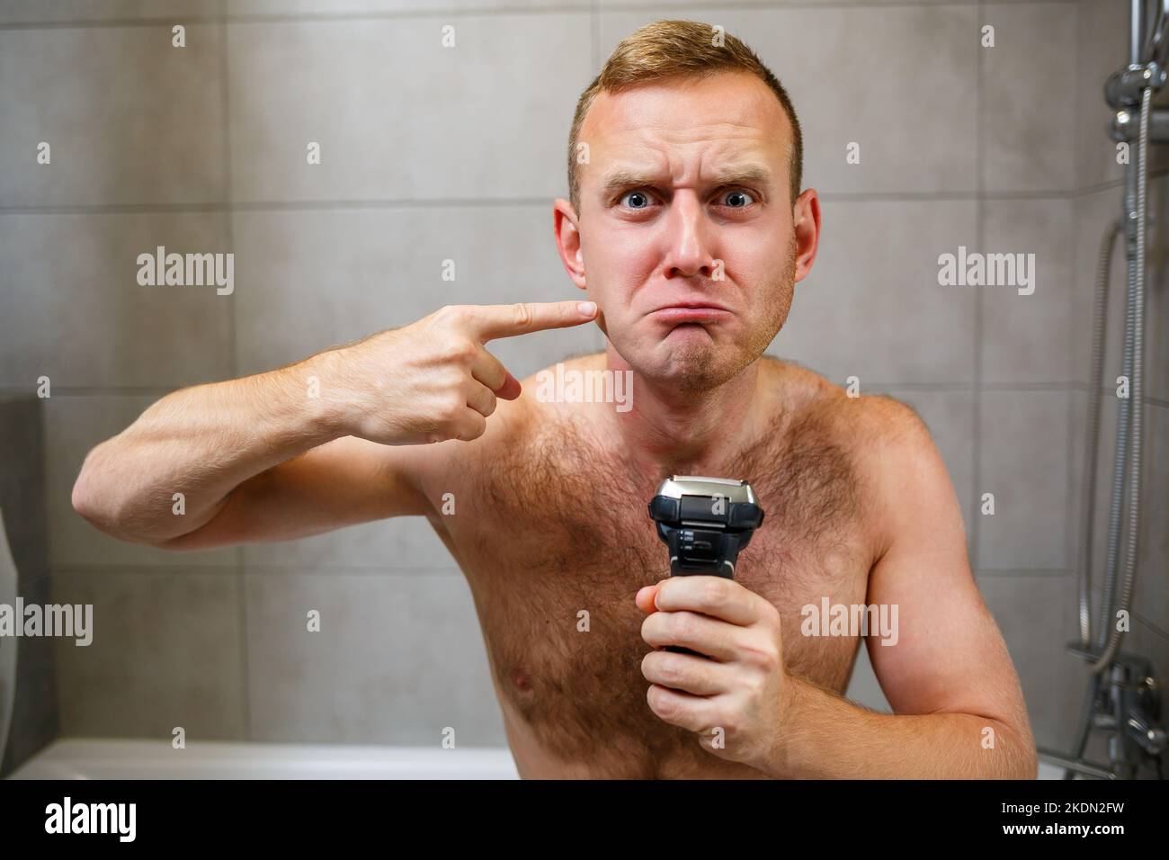 A man shaves his face with an electric razor in front of a mirror. Skin ...