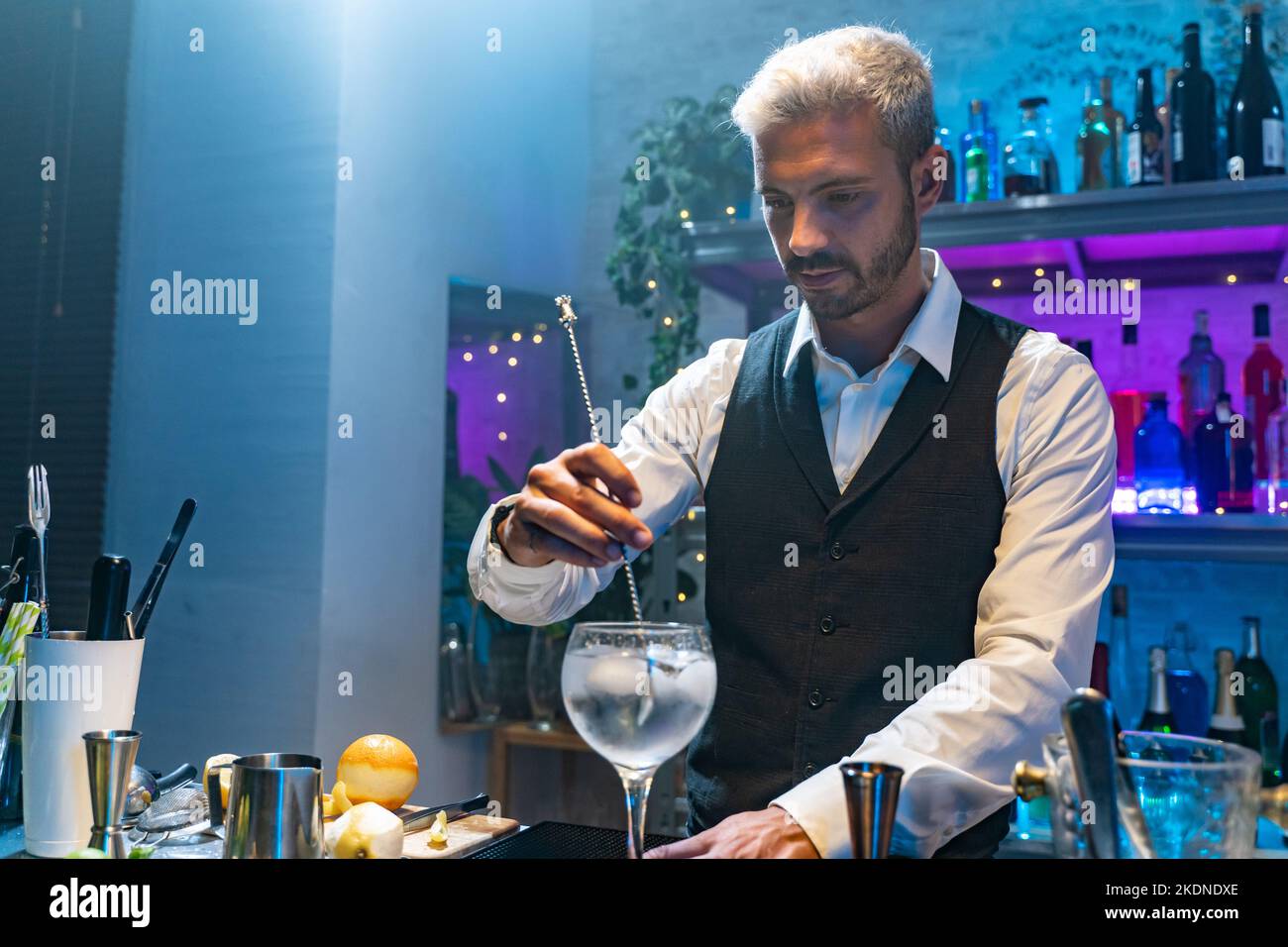 Young barman in a white shirt and black apron making cocktail at party ...