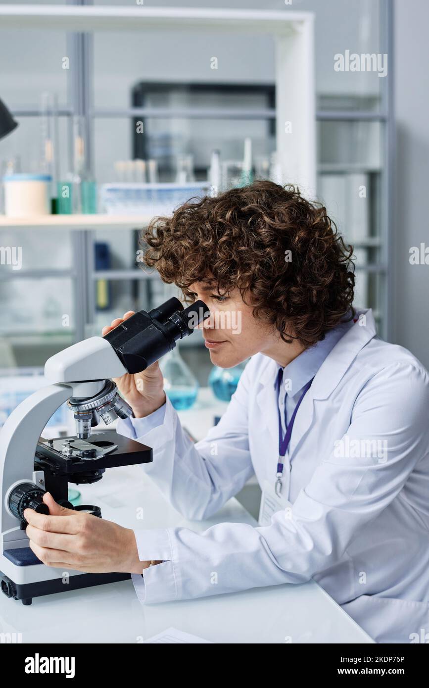 Young serious female researcher in labcoat looking in microscope while ...