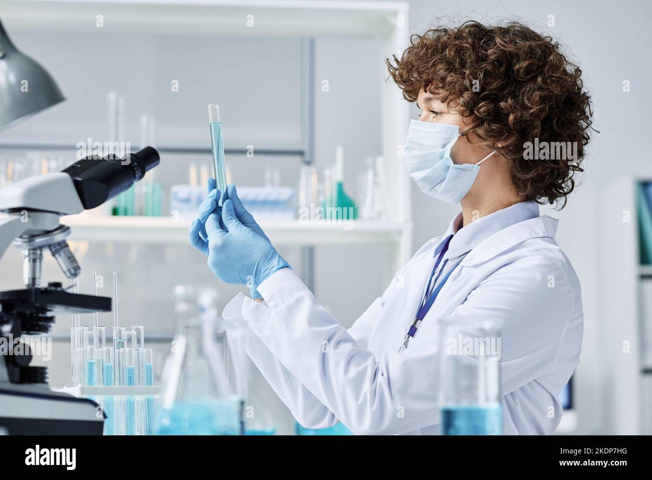 Young virologist in protective mask and gloves holding flask with blue ...