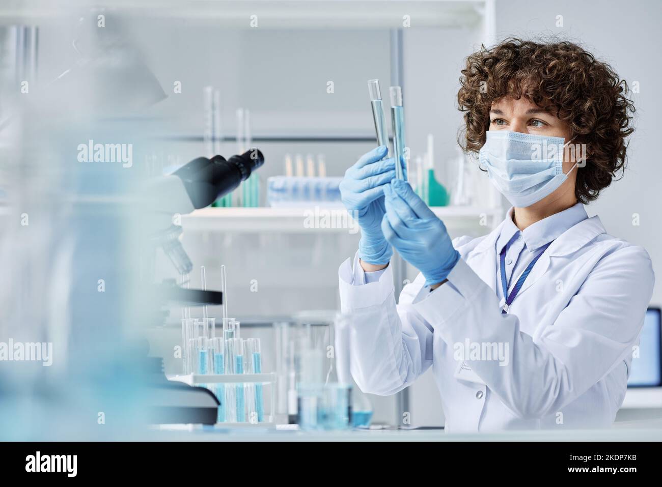 Young female virologist in protective workwear looking at two flasks ...