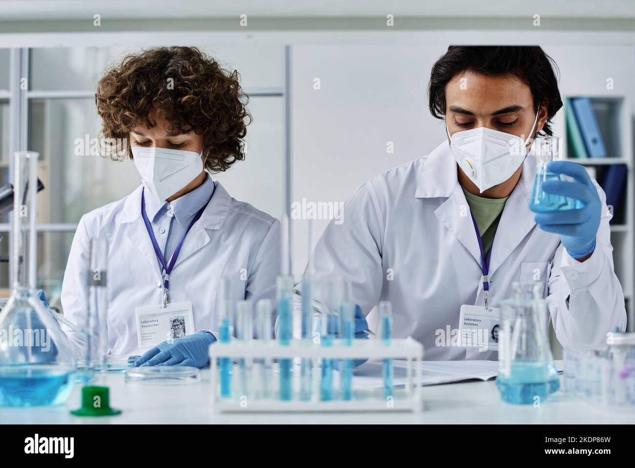 Two young clinicians in labcoats, respirators and gloves working with ...