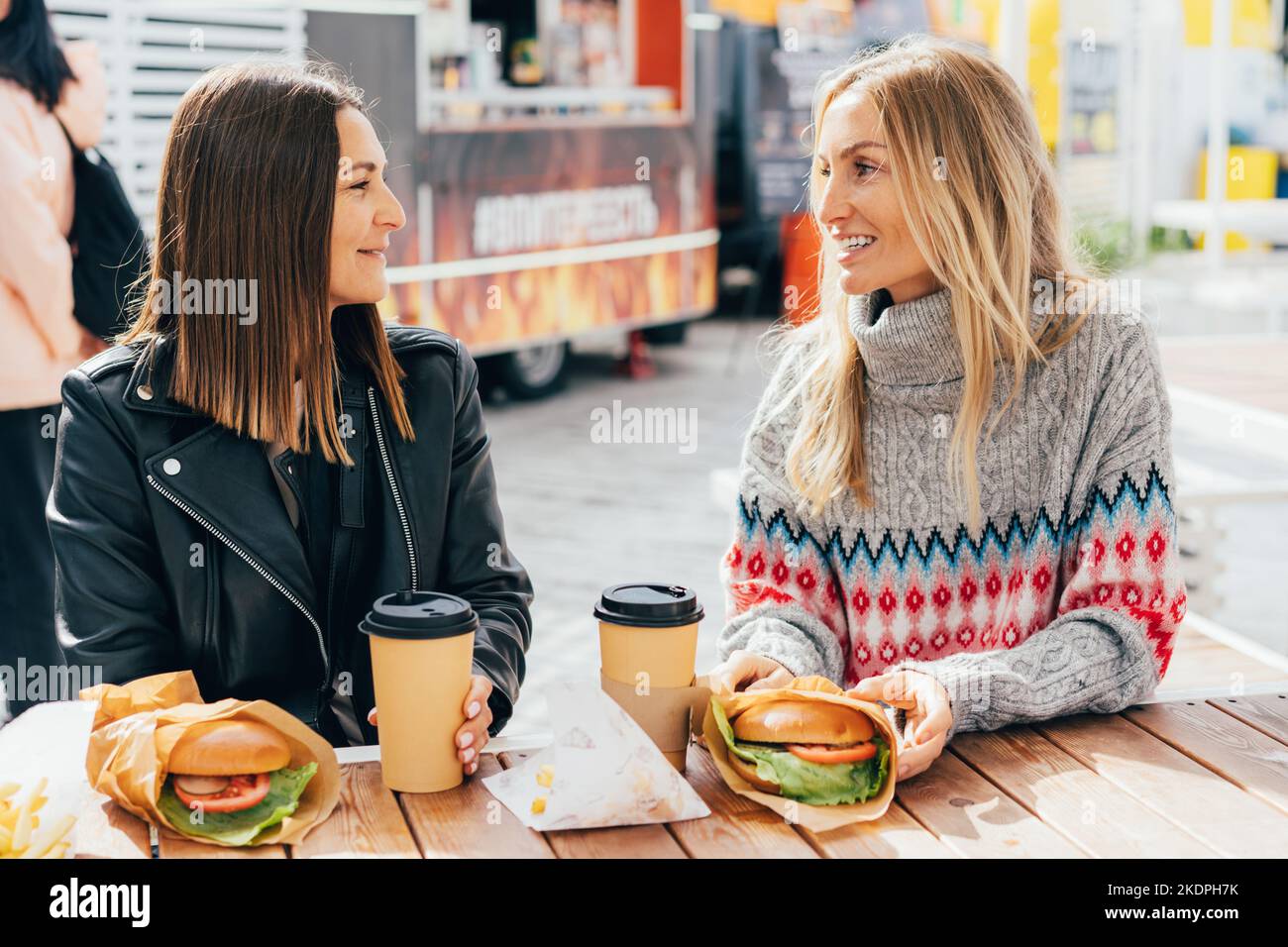 Two happy hungry female friends are chatting and eating burgers at the ...