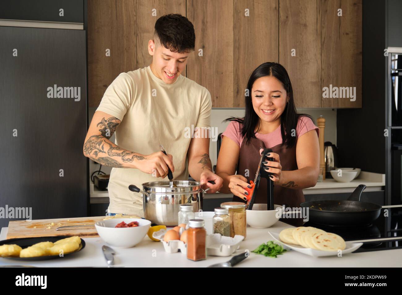 Two young latin people mashing potatoes and grating tomato at kitchen ...