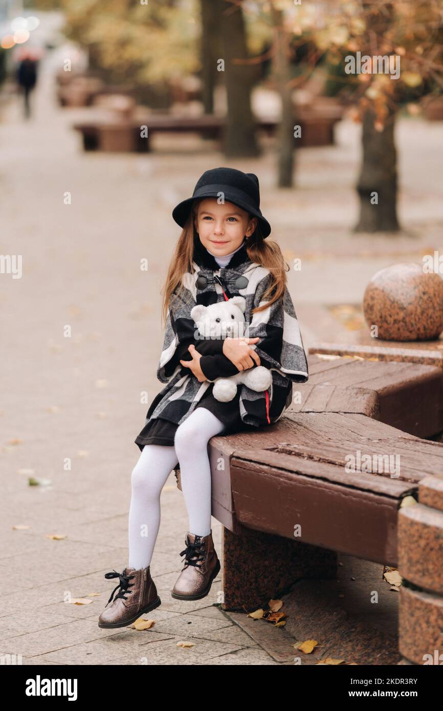 A little stylish girl in autumn clothes is sitting on a bench in the ...