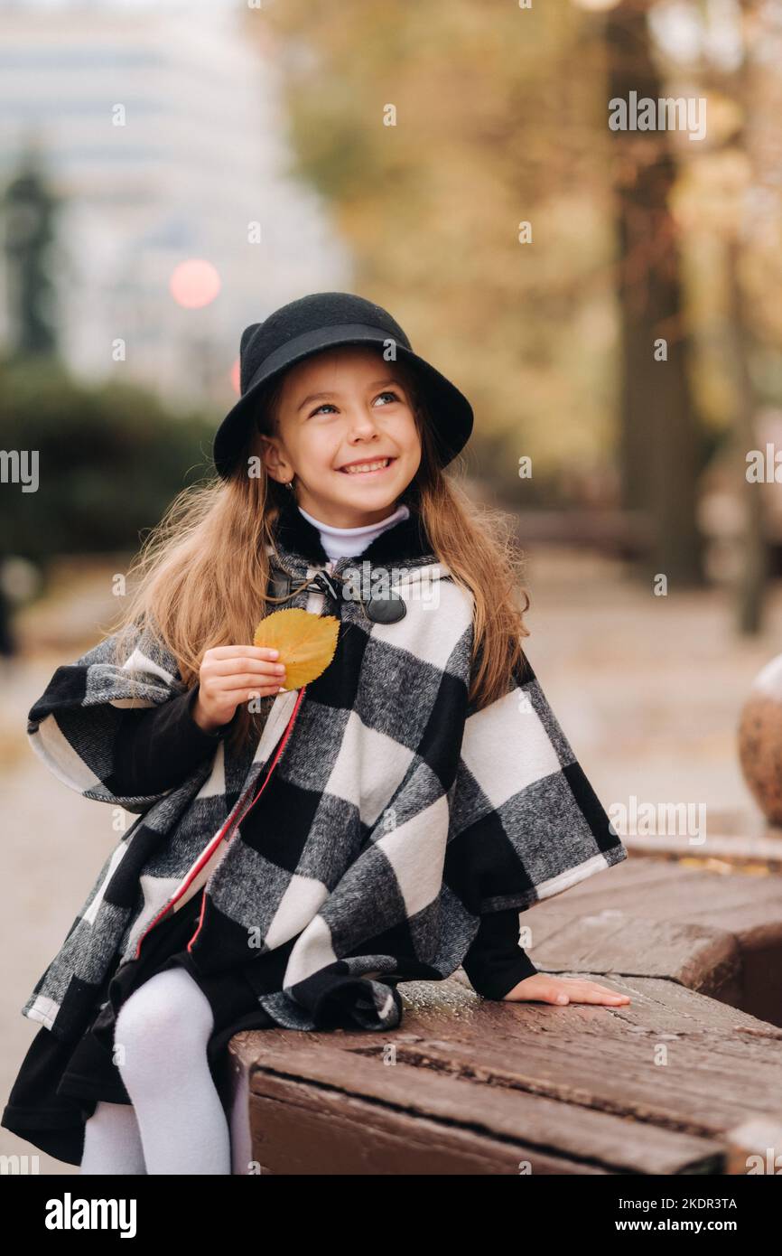 A little stylish girl in autumn clothes is sitting on a bench in the ...