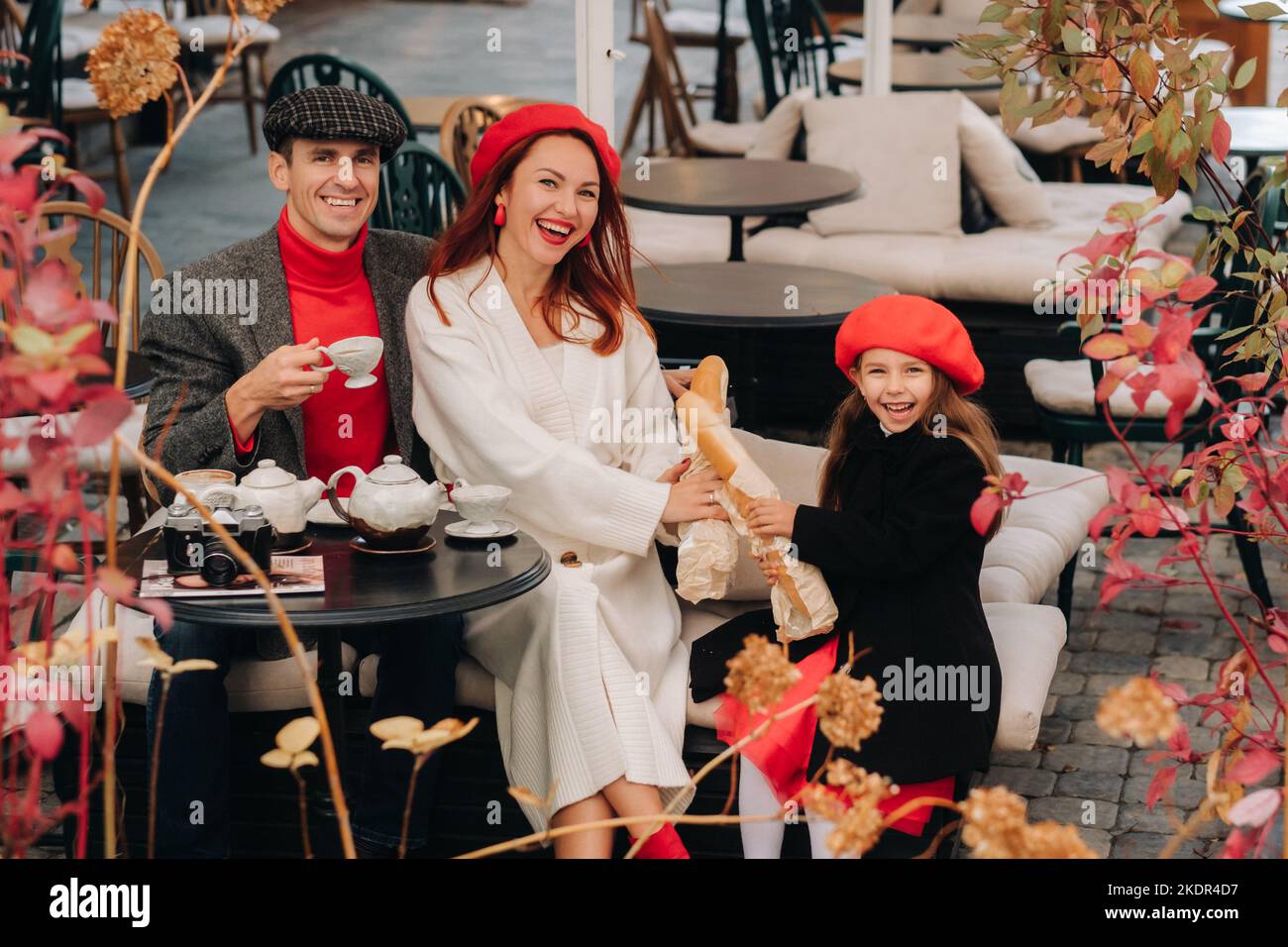 A stylish family of three is sitting at a table outside in a cafe and ...