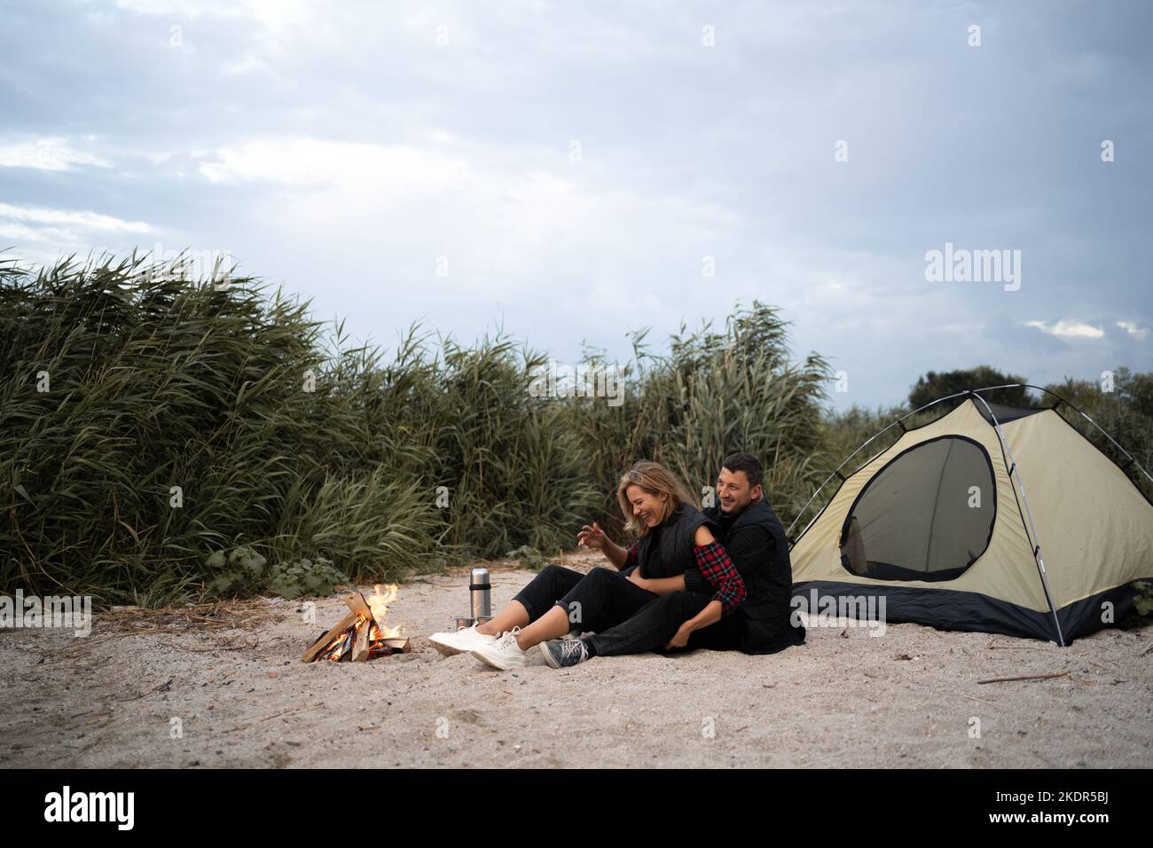 Romantic couple tourists laugh sitting at a campfire near tent, hugging ...