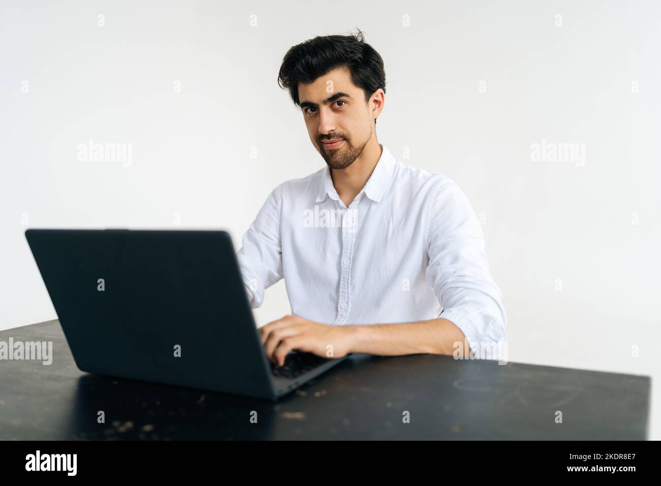 Studio portrait of friendly young man in shirt working on laptop ...