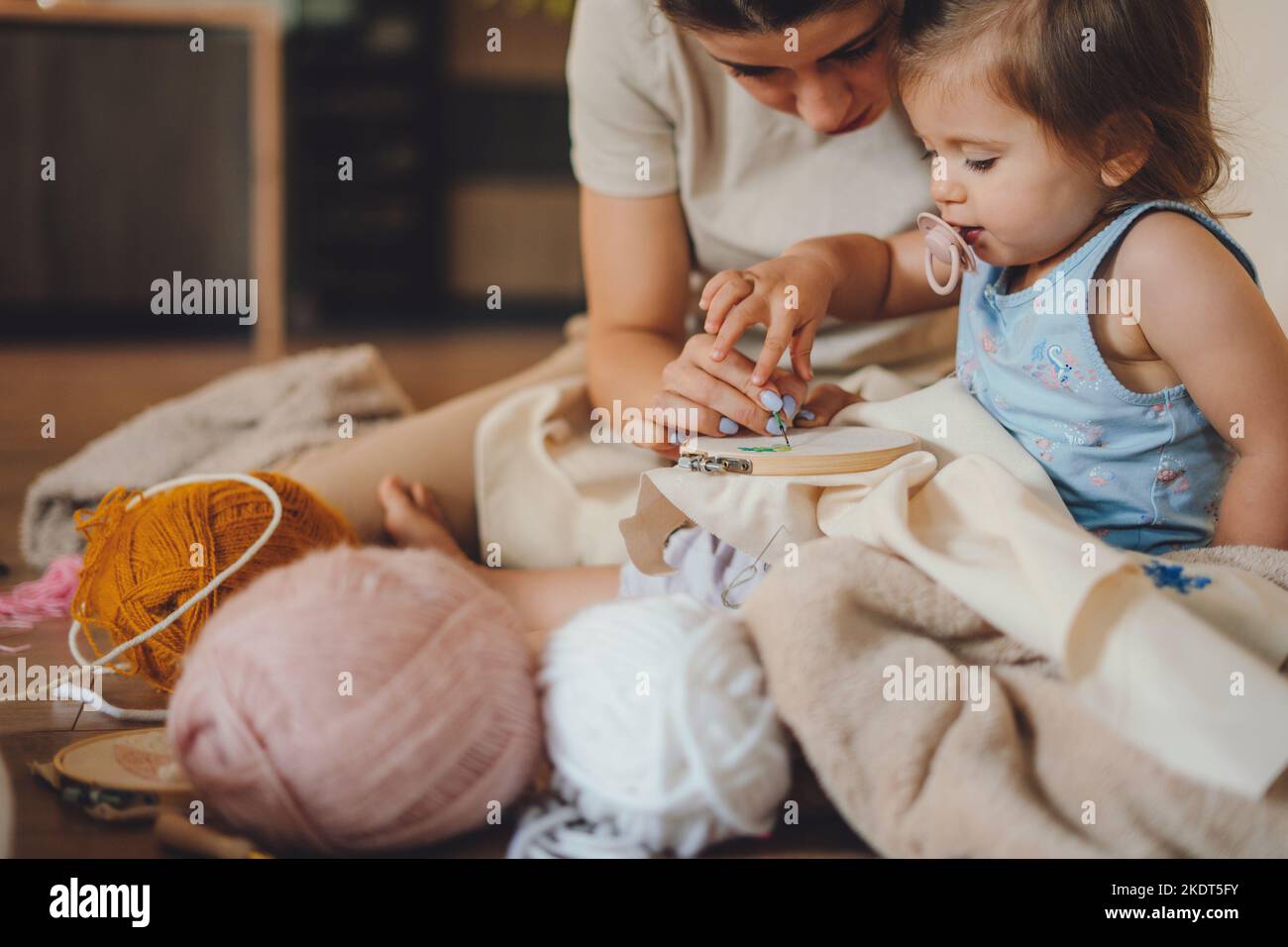 The caring mother teaching her child to embroider from an early age sitting on the floor. Hobby ...